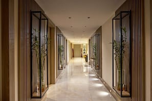 Um corredor com piso de mármore polido, ladeado por painéis verticais de madeira e vitrines de vidro com plantas.