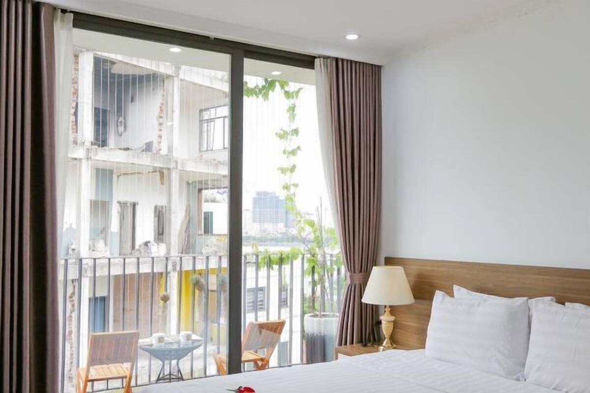 Quarto de hotel com cama, mesa de cabeceira, luminária, janela com cortinas e vista para edifícios.