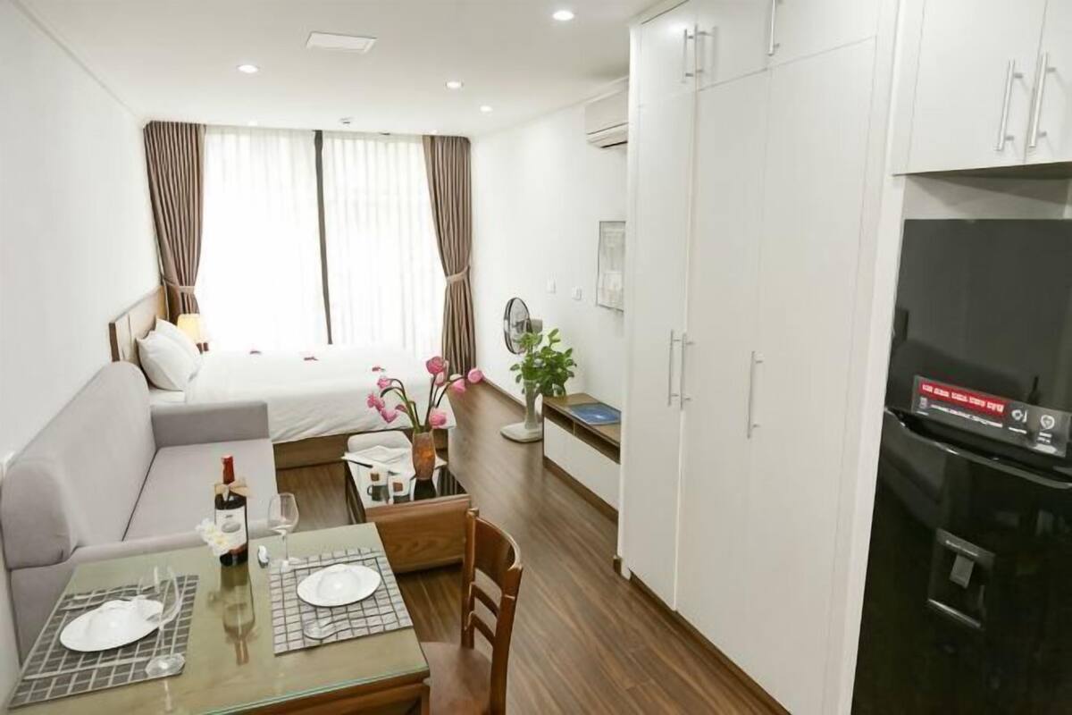 Quarto de hotel compacto com cama, mesa de jantar com cadeiras, pequena cozinha com geladeira e janela com cortinas.