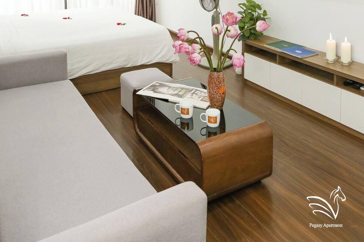 Quarto de hotel moderno com cama, sofá, mesa de centro com um vaso de flores, TV fixada na parede e aparelho de ar condicionado.