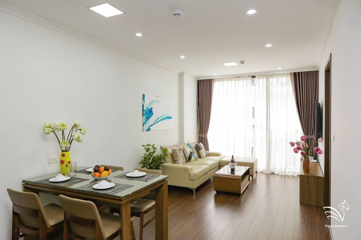 Uma sala de estar moderna com área de jantar, um sofá e uma mesa de centro. Há um quadro na parede e uma janela com cortinas.