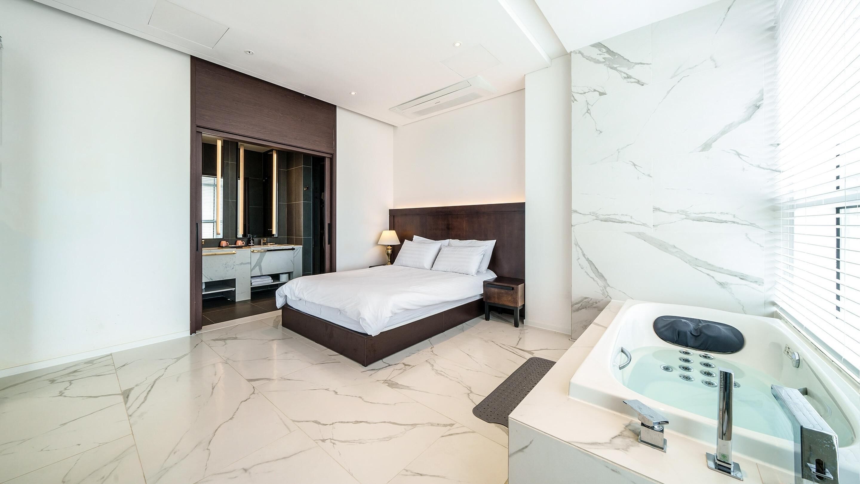 Quarto de hotel moderno com uma cama grande, banheiro separado com hidromassagem, piso de mármore e cabeceira de madeira escura.