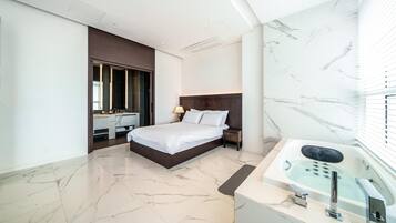 Quarto de hotel moderno com uma cama grande, banheiro separado com hidromassagem, piso de mármore e cabeceira de madeira escura.