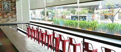 Uma área de estar com cadeiras vermelhas e um banco comprido, grandes janelas com vista para a rua e vegetação, e uma parede de tijolos com um quadro emoldurado.