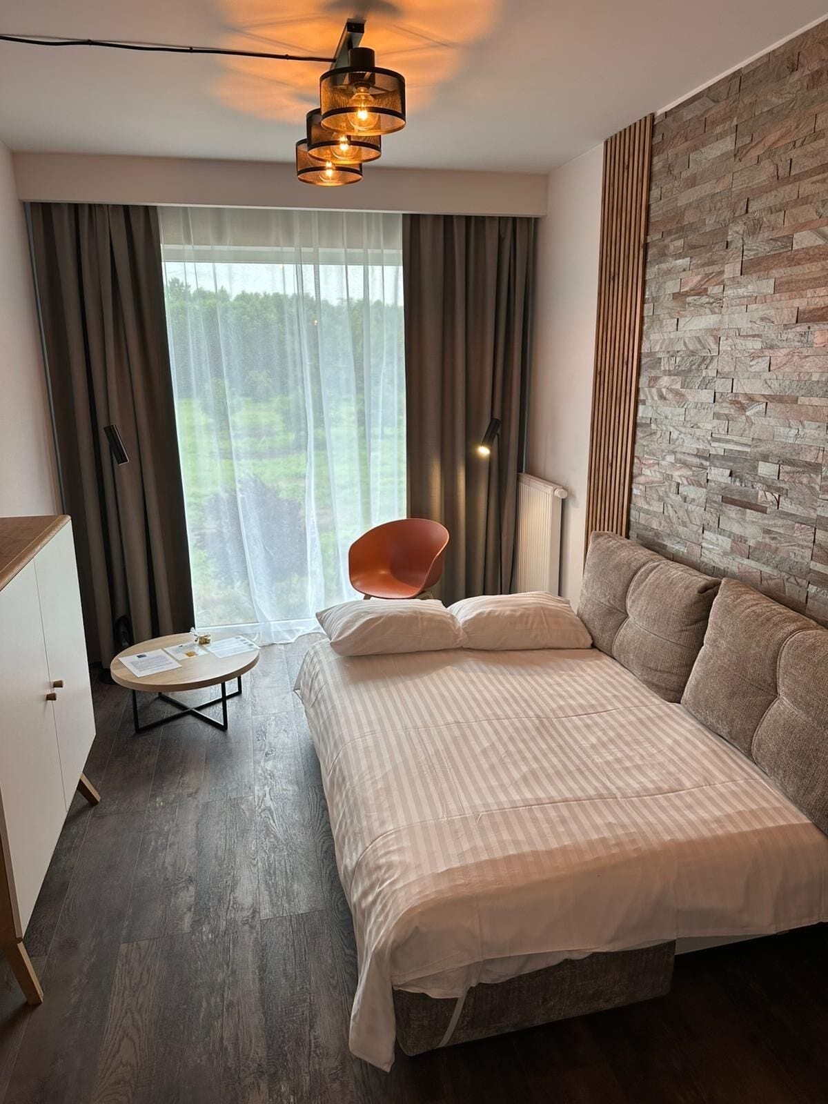 Quarto moderno com cama, cadeira, mesinha de apoio e vista para o verde pela janela.