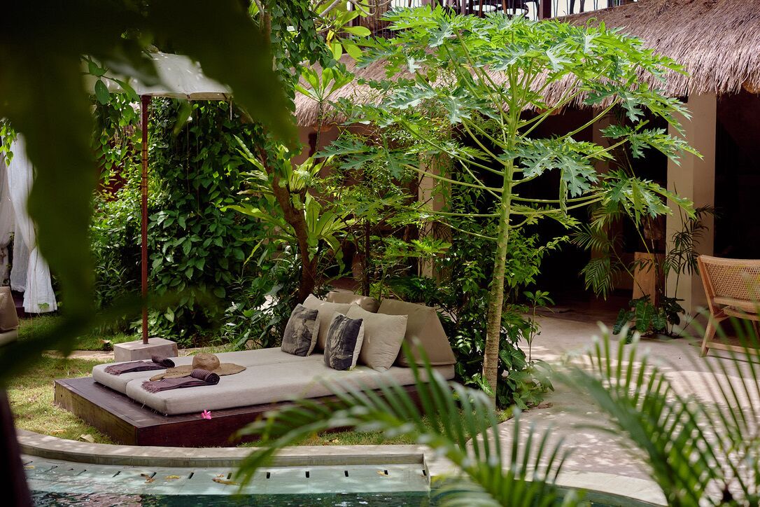 Uma área junto à piscina com espreguiçadeira e almofadas, cercada por vegetação exuberante e uma estrutura com telhado de palha.