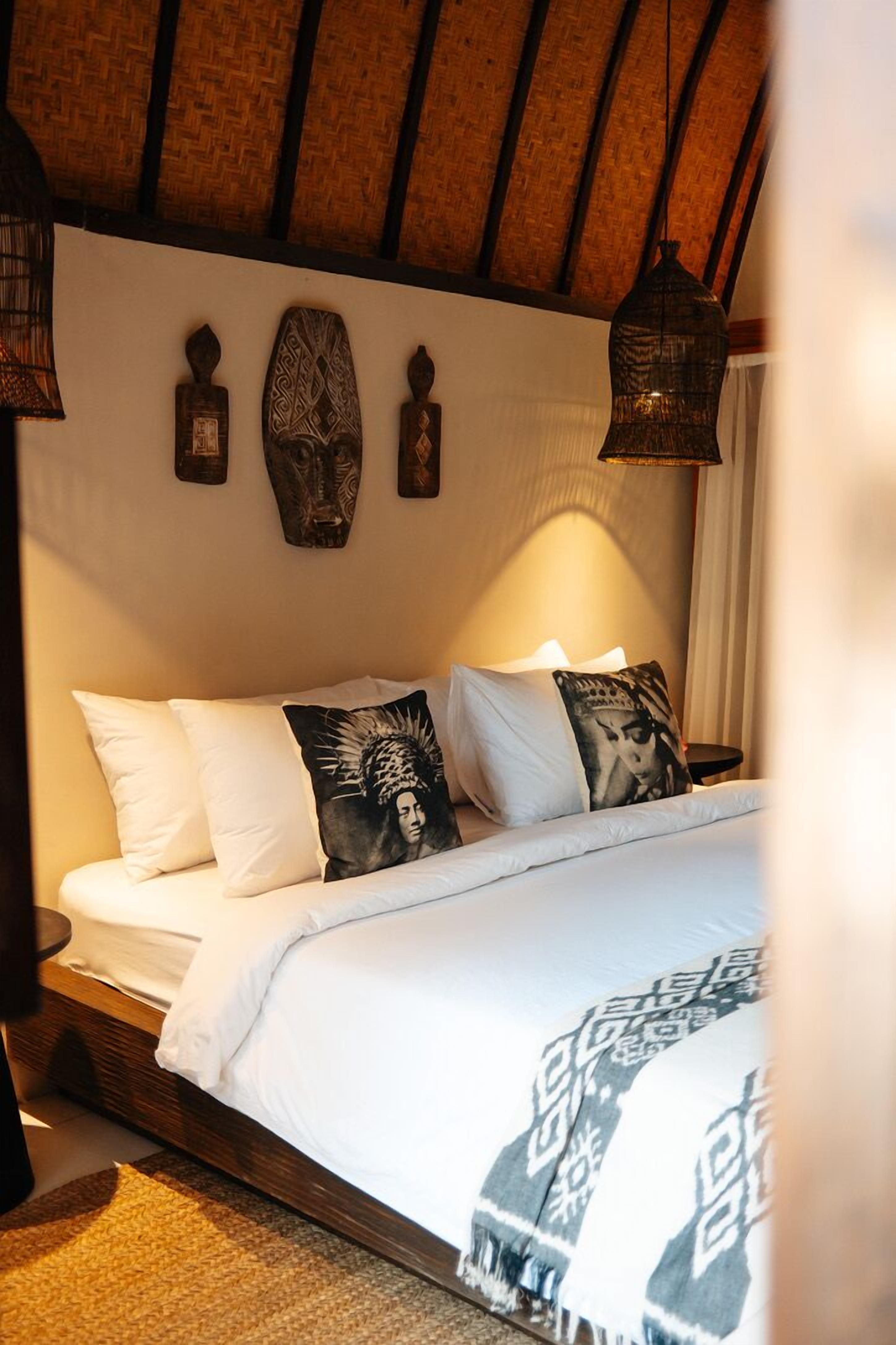 Cama com roupa de cama branca e almofadas estampadas, tapete tecido e teto com vigas de madeira.