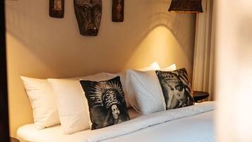 Cama com roupa de cama branca e almofadas estampadas, tapete tecido e teto com vigas de madeira.