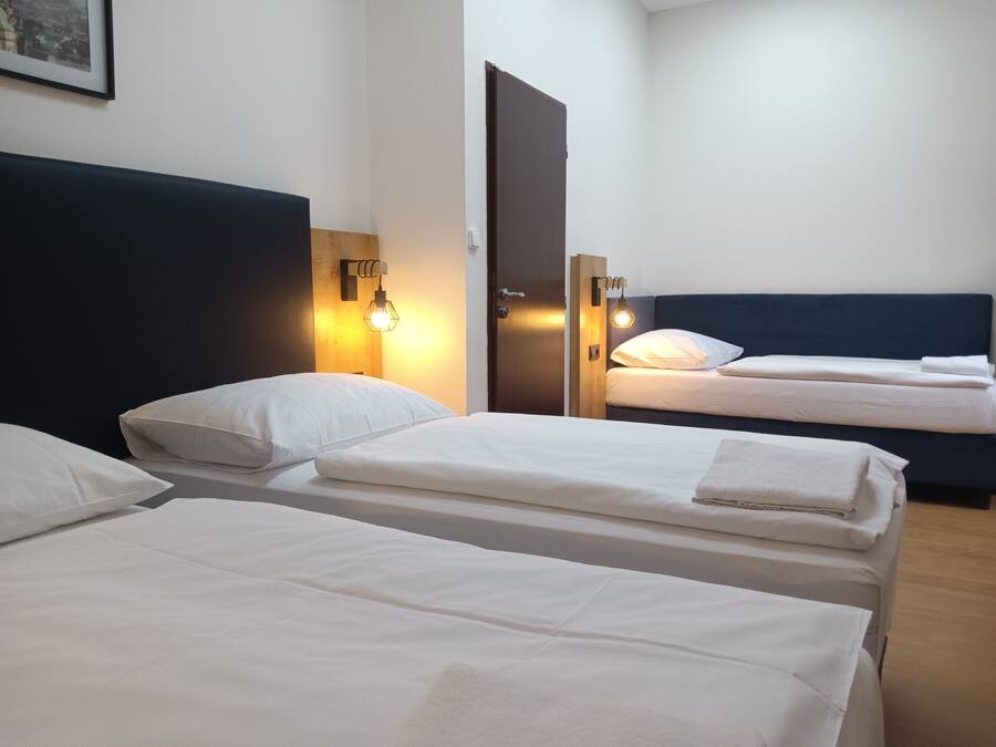 Quarto de hotel com duas camas de solteiro, cabeceira escura, porta de madeira e luminárias fixadas na parede.