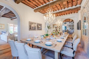Uma sala de jantar com uma mesa comprida posta para refeição, rodeada por cadeiras estofadas.