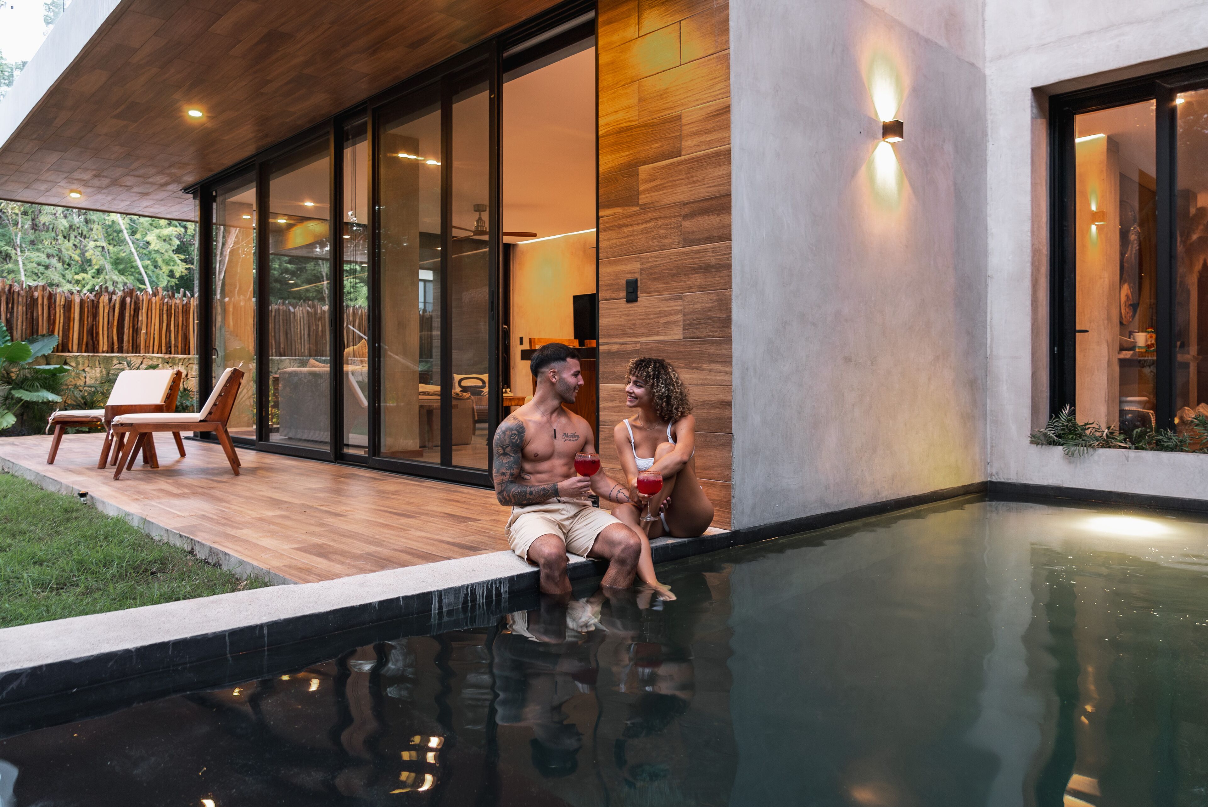 Área moderna de piscina ao ar livre com estrutura de vidro, deck de madeira e espreguiçadeiras.