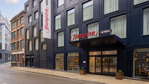Um hotel moderno com um destaque para a placa 'Hampton by Hilton', grandes janelas e fachada limpa.