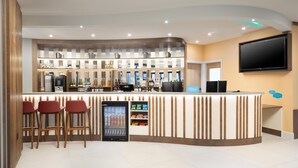 Uma área de recepção de hotel com balcão, banquetas, dispensador de bebidas e uma televisão na parede.