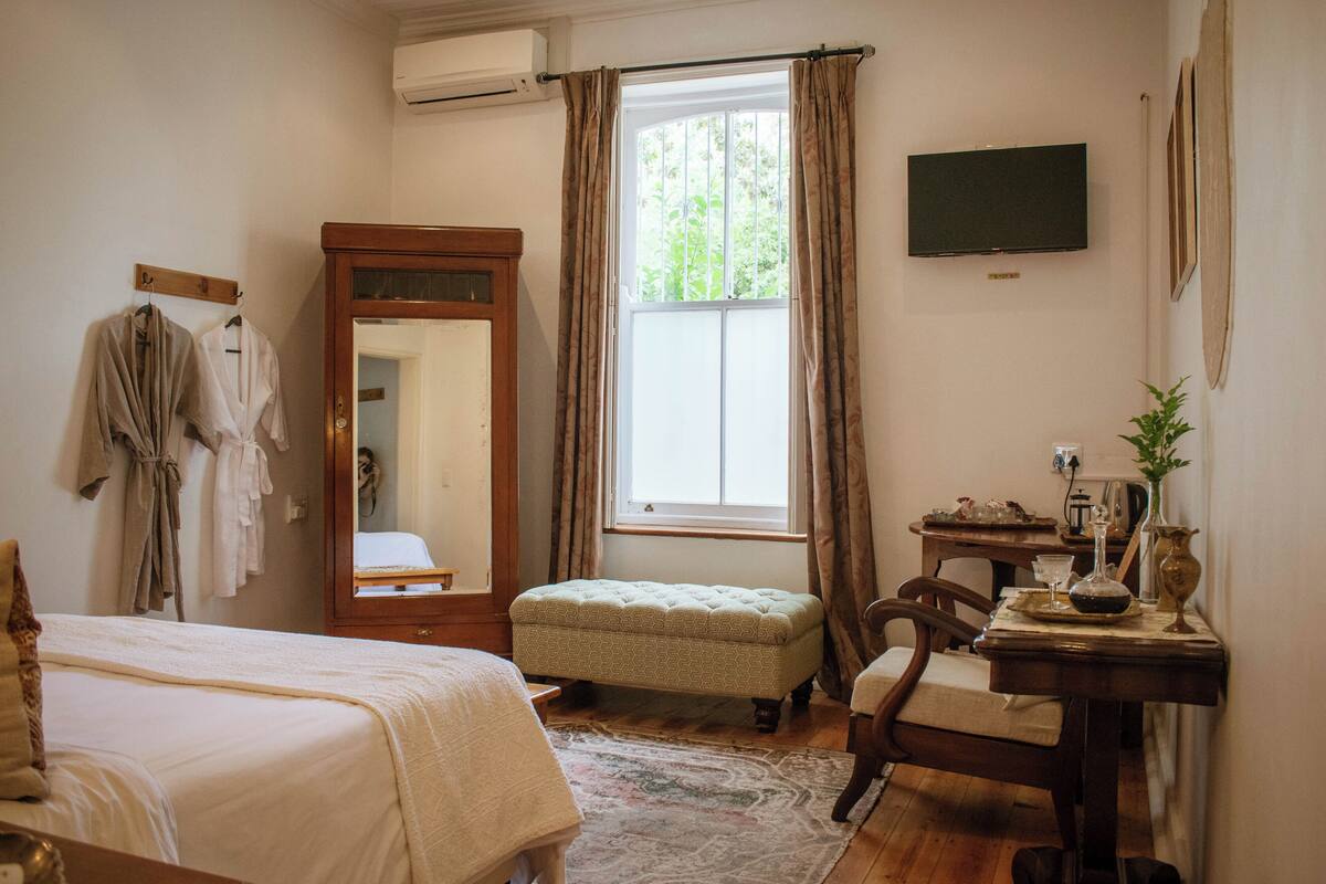 Quarto com uma cama, um banco de madeira, uma mesa com um vaso e um espelho.
