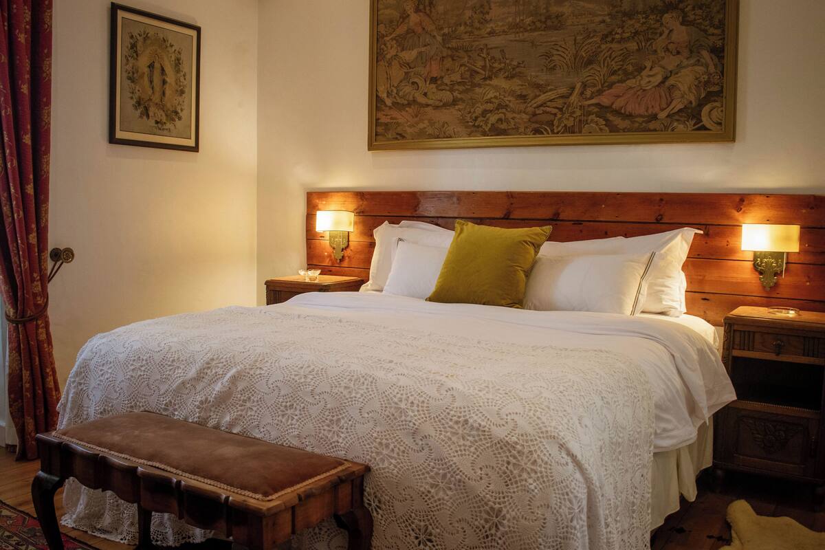 Uma cama bem arrumada com cabeceira de madeira, duas luminárias de apoio e um banquinho no rodapé da cama.