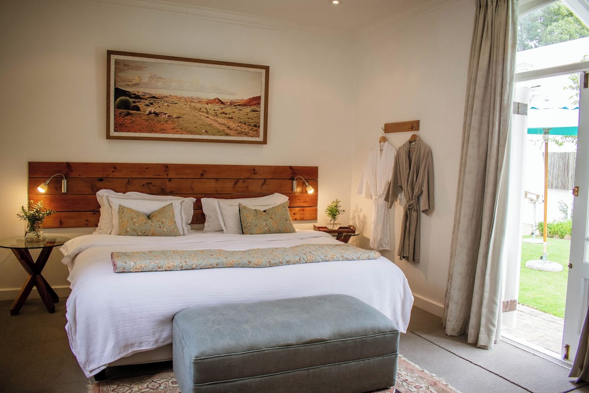 Quarto com uma cama grande, cabeceira de madeira, pintura sobre a cama, mesa de cabeceira com abajur e vista para o exterior através de uma porta.