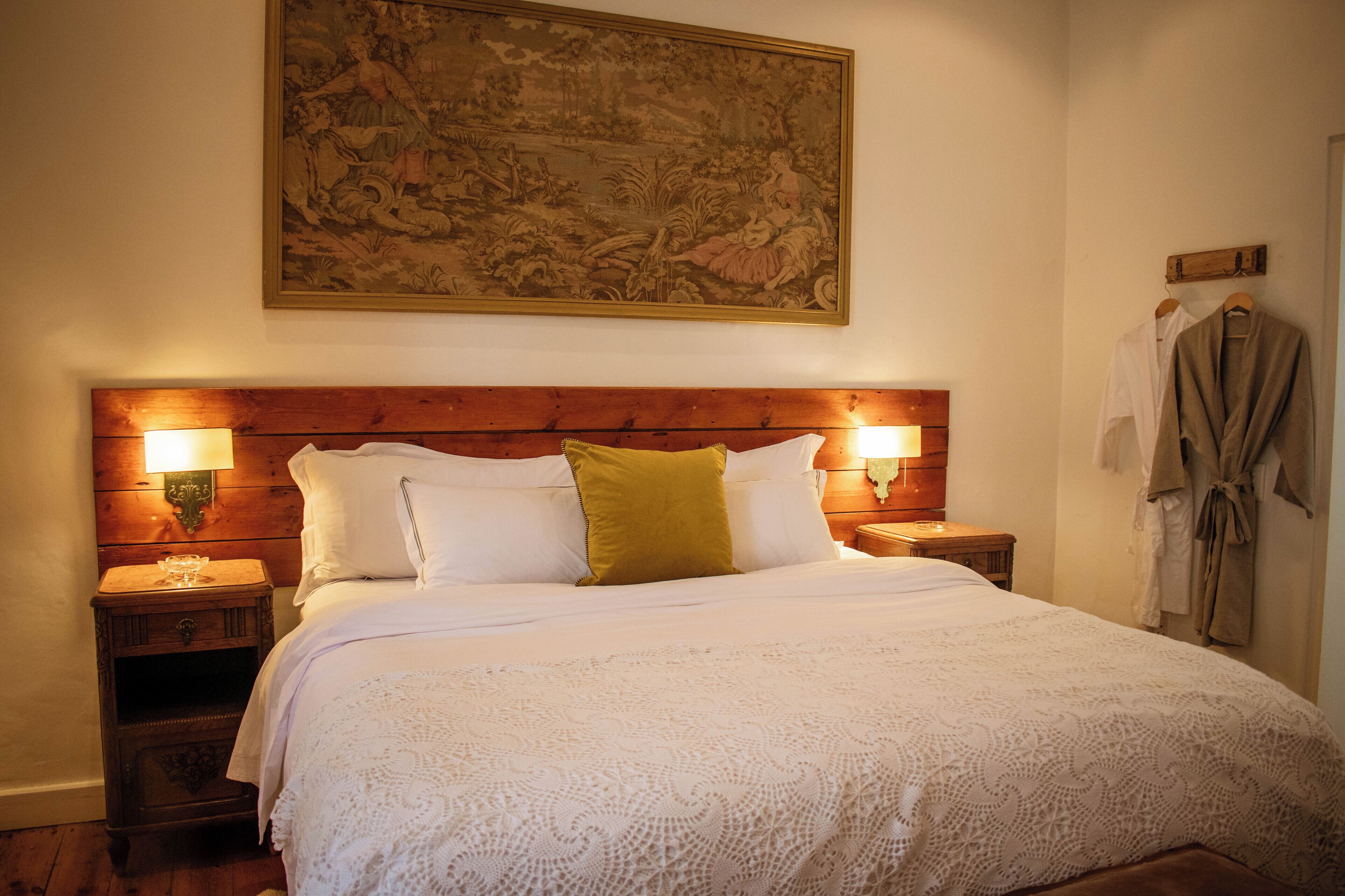 Cama bem arrumada com almofadas brancas e amarelas, cabeceira de madeira e um quadro acima.