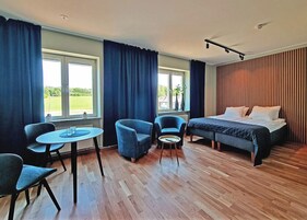 Quarto de hotel moderno com uma cama, duas poltronas azuis, uma mesa redonda pequena e vista para o exterior.