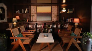 Uma área de estar aconchegante, com cadeiras de madeira e uma mesa central, cercada por estantes de livros e objetos decorativos.