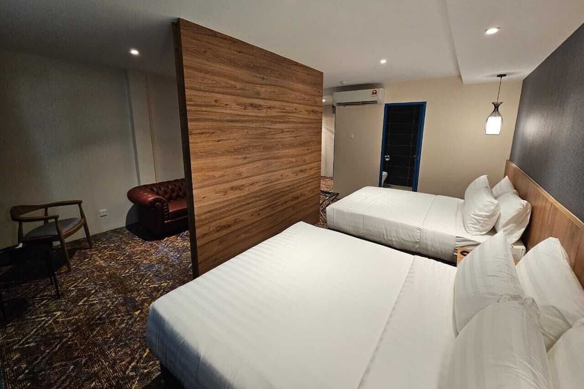 Quarto de hotel com duas camas, divisória de madeira, sofá de couro vermelho, tapete estampado e luminária no teto.