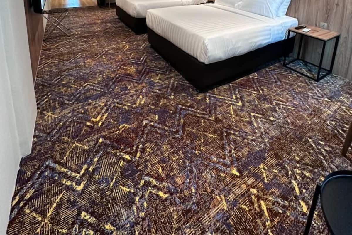 Quarto de hotel com duas camas, um tapete estampado, uma parede texturizada cinza e uma mesinha de cabeceira.