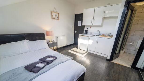 Quarto de hotel moderno com uma cama bem arrumada, uma pequena mesa de cabeceira com abajur, uma cozinha compacta e uma porta para um banheiro.