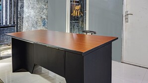 Uma mesa de escritório moderna com acabamento escuro e tampo de madeira, posicionada em um ambiente com piso de azulejos e uma porta ao fundo.