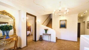 Um hall imponente com um espelho grande e ricamente decorado, um candelabro e uma escadaria que leva a outro cômodo.
