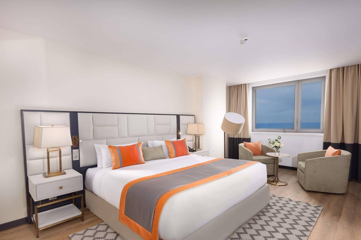 Quarto de hotel moderno com uma cama grande, mesas de cabeceira, escrivaninha e vista para o mar.