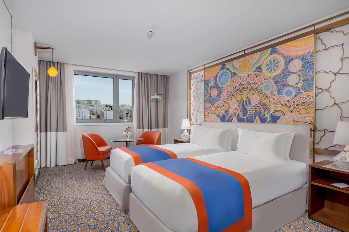 Quarto de hotel com uma cama grande, duas mesas de cabeceira, uma escrivaninha, uma cadeira, uma televisão e uma janela com cortinas.