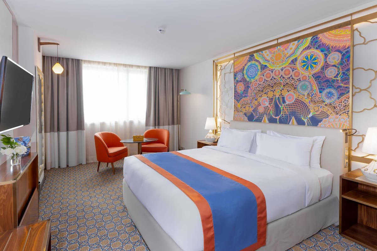 Quarto de hotel com uma cama grande, duas cadeiras laranja, uma mesa pequena, uma televisão e um mural colorido na parede.