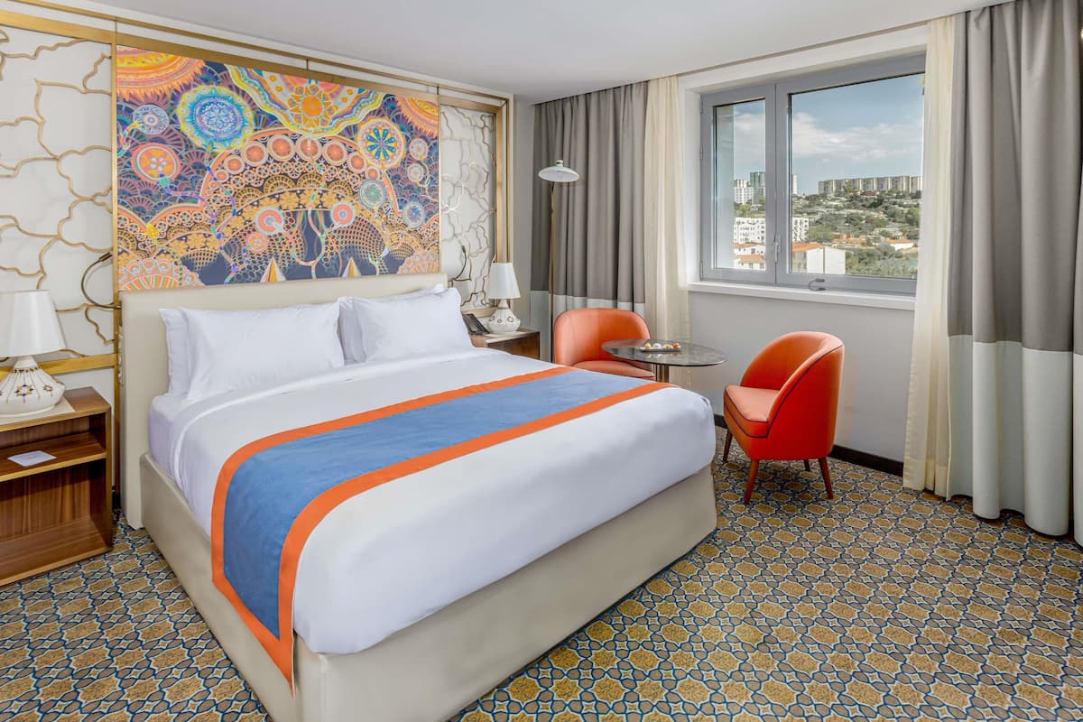 Quarto de hotel com uma cama grande, uma escrivaninha com cadeira, uma janela com vista para a cidade e um quadro abstrato colorido na parede.