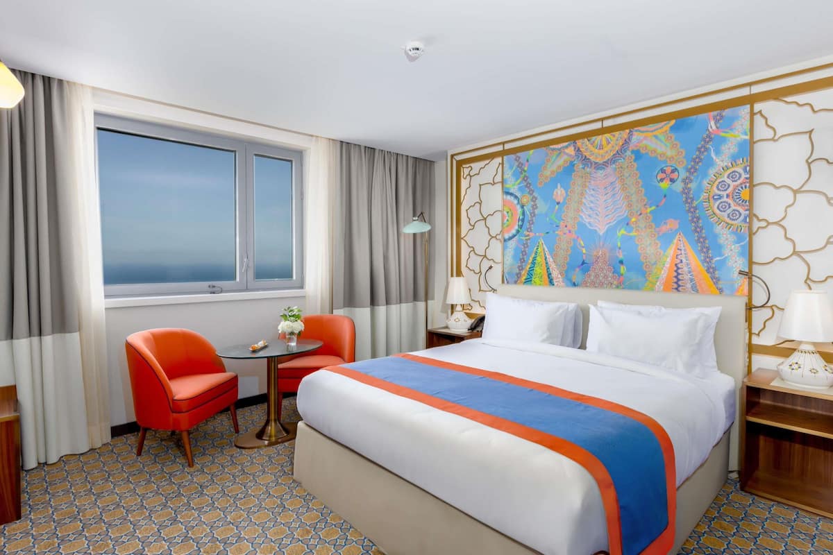 Quarto de hotel com uma cama grande, uma mesa pequena, duas cadeiras, uma mesa de cabeceira e uma janela com cortinas.
