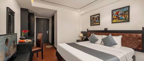 Quarto de hotel com cama, escrivaninha com computador, cadeira, televisão e obras de arte nas paredes.
