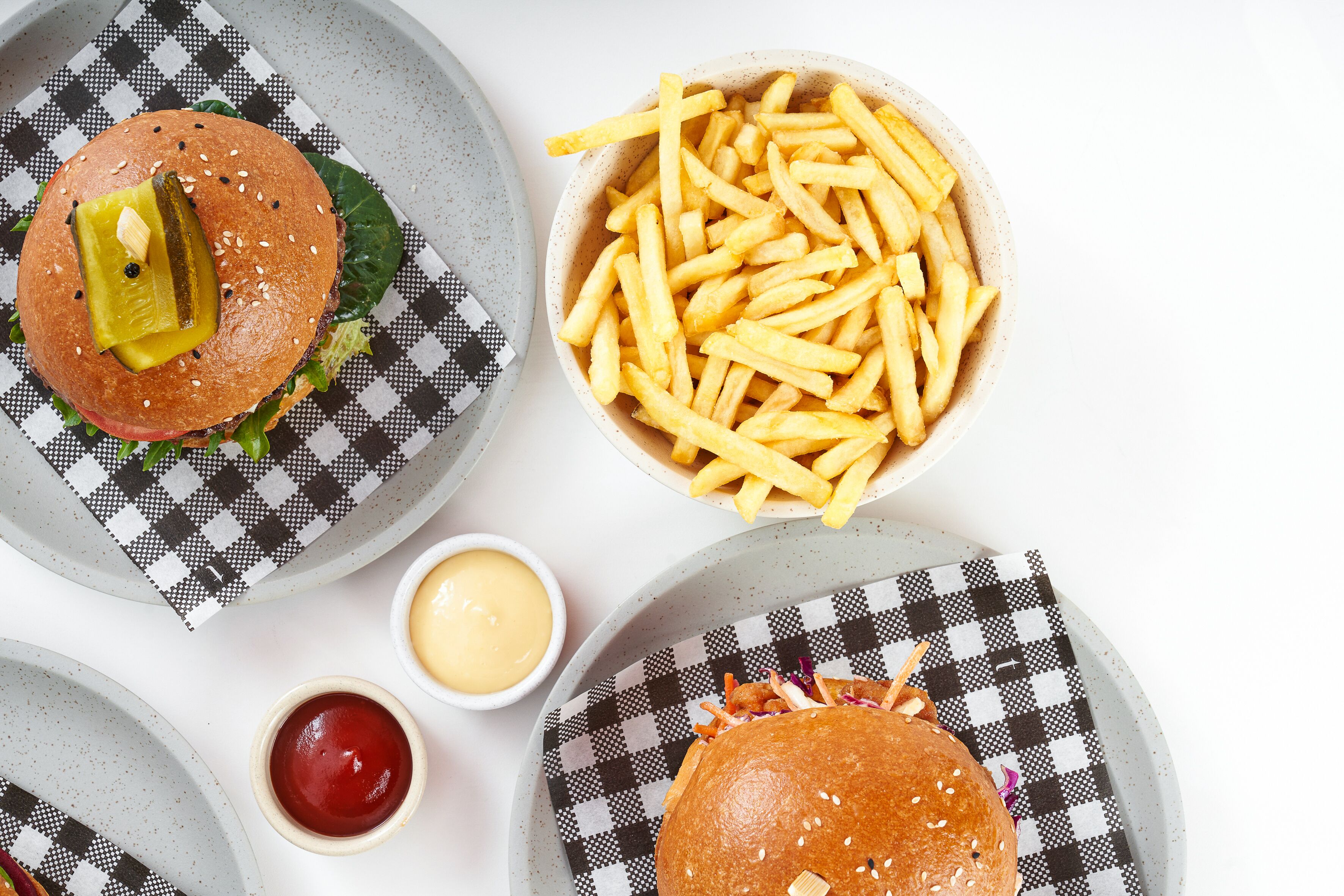 Uma refeição com hambúrguer, picles, batatas fritas e dois acompanhamentos em uma toalha de mesa xadrez.