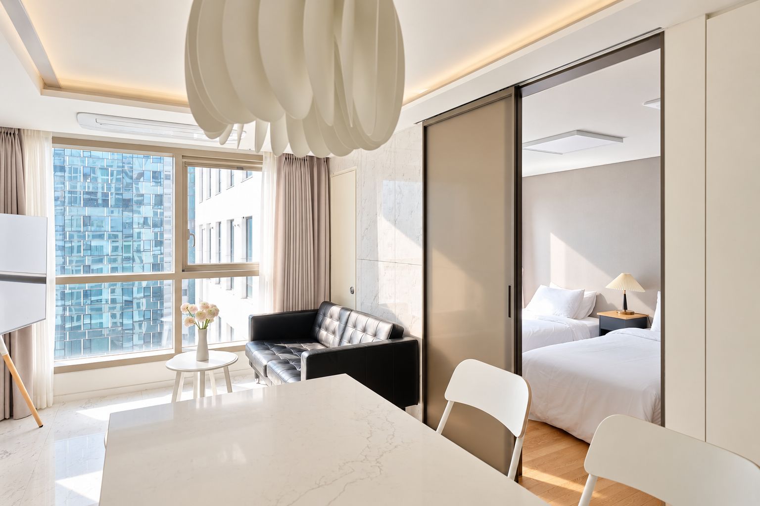 Quarto de hotel moderno com uma cama grande, sofá, mesa de jantar e vista para a cidade através de janelas do chão ao teto.
