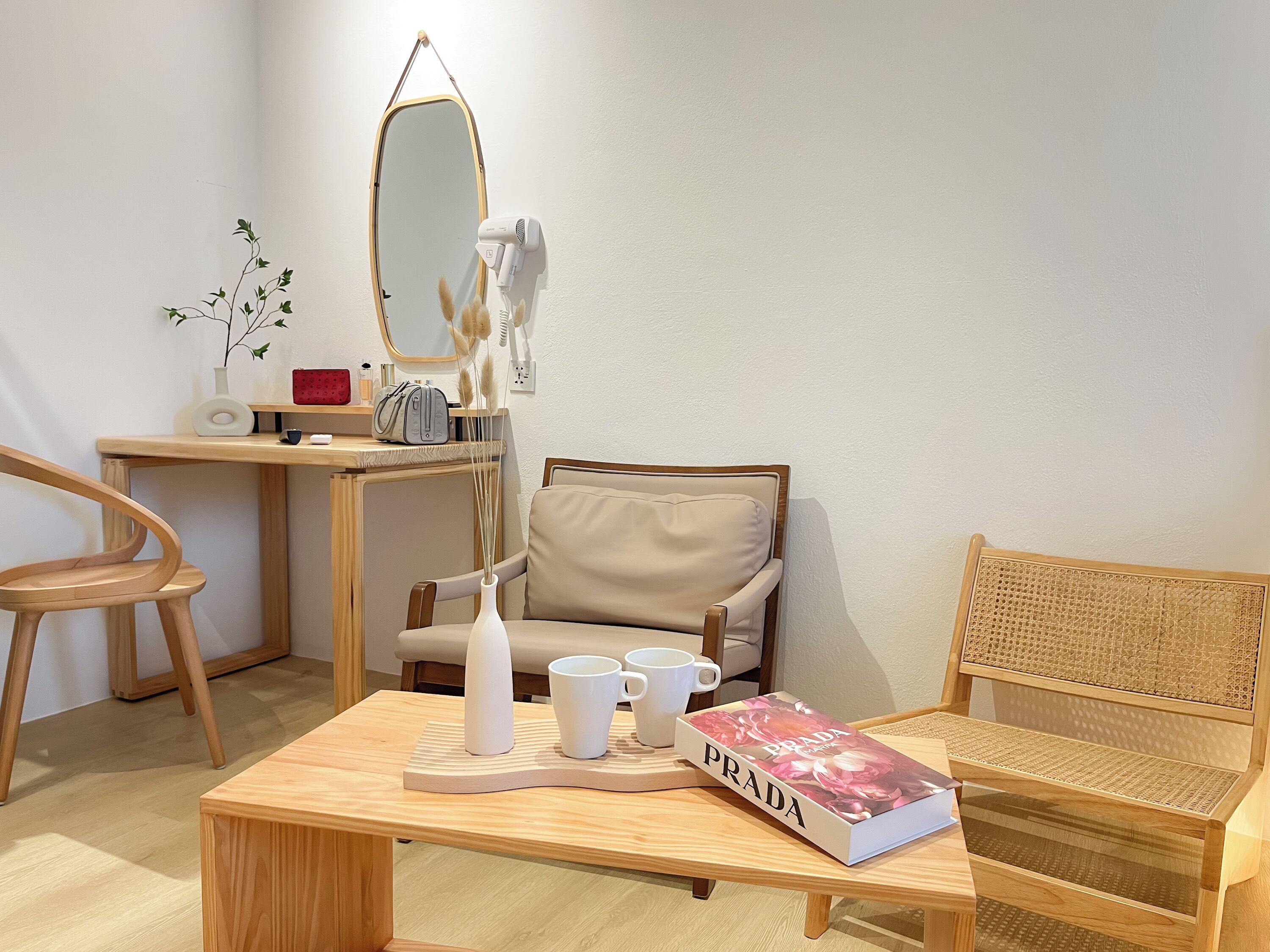 Quarto com escrivaninha de madeira, uma cadeira, toucador com espelho, secador de cabelo, vaso com plantas secas, um livro intitulado 'PRADA' e duas canecas brancas.