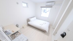 Quarto moderno e minimalista com cama, escrivaninha e cadeira. Há uma janela que oferece luz natural e uma porta com fechadura eletrônica.