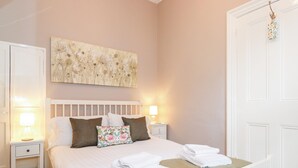 Cama arrumada com almofadas e manta floral, luminária de cabeceira e obra de arte na parede em quarto de hotel.