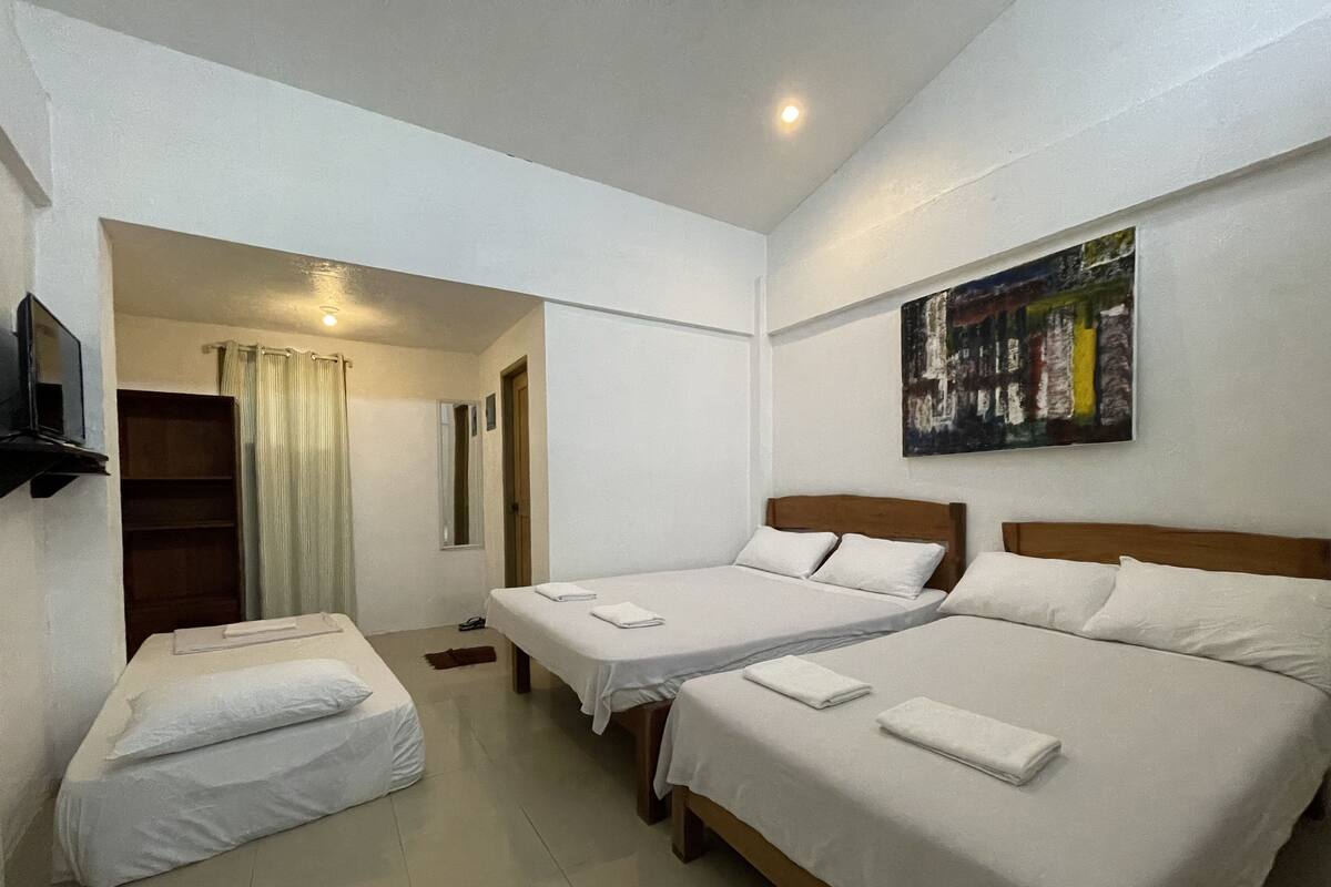 Quarto de hotel com duas camas de solteiro e uma cama-sofá, uma TV fixada na parede e um quadro sobre as camas.