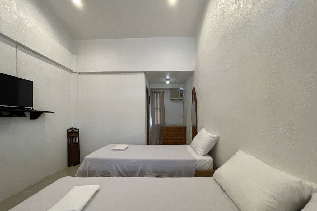 Quarto pequeno e bem iluminado com duas camas, uma televisão de tela plana na parede e um aparador de madeira.
