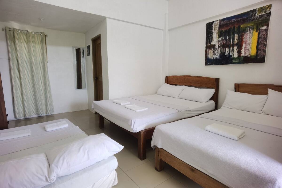 Quarto de hotel com três camas, uma grande pintura abstrata na parede e uma janela com cortinas.