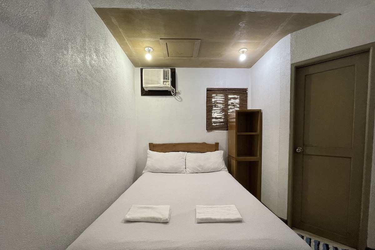 Uma cama pequena e bem arrumada, com lençóis brancos e duas toalhas dobradas, um cabeceira de madeira, um ar condicionado fixado na parede, uma porta e uma janela com persianas.