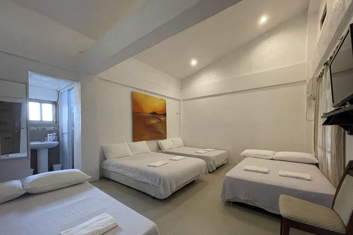 Quarto de hotel com três camas de solteiro, área de estar com uma poltrona, um quadro na parede e banheiro com pia e espelho.
