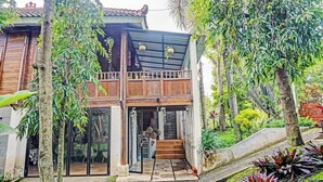 Uma casa de madeira com varanda, rodeada por vegetação exuberante e um pequeno espelho d'água.