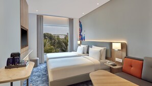 Quarto de hotel com duas camas, uma escrivaninha, uma cadeira, televisão e janela com vista para árvores e edifícios.