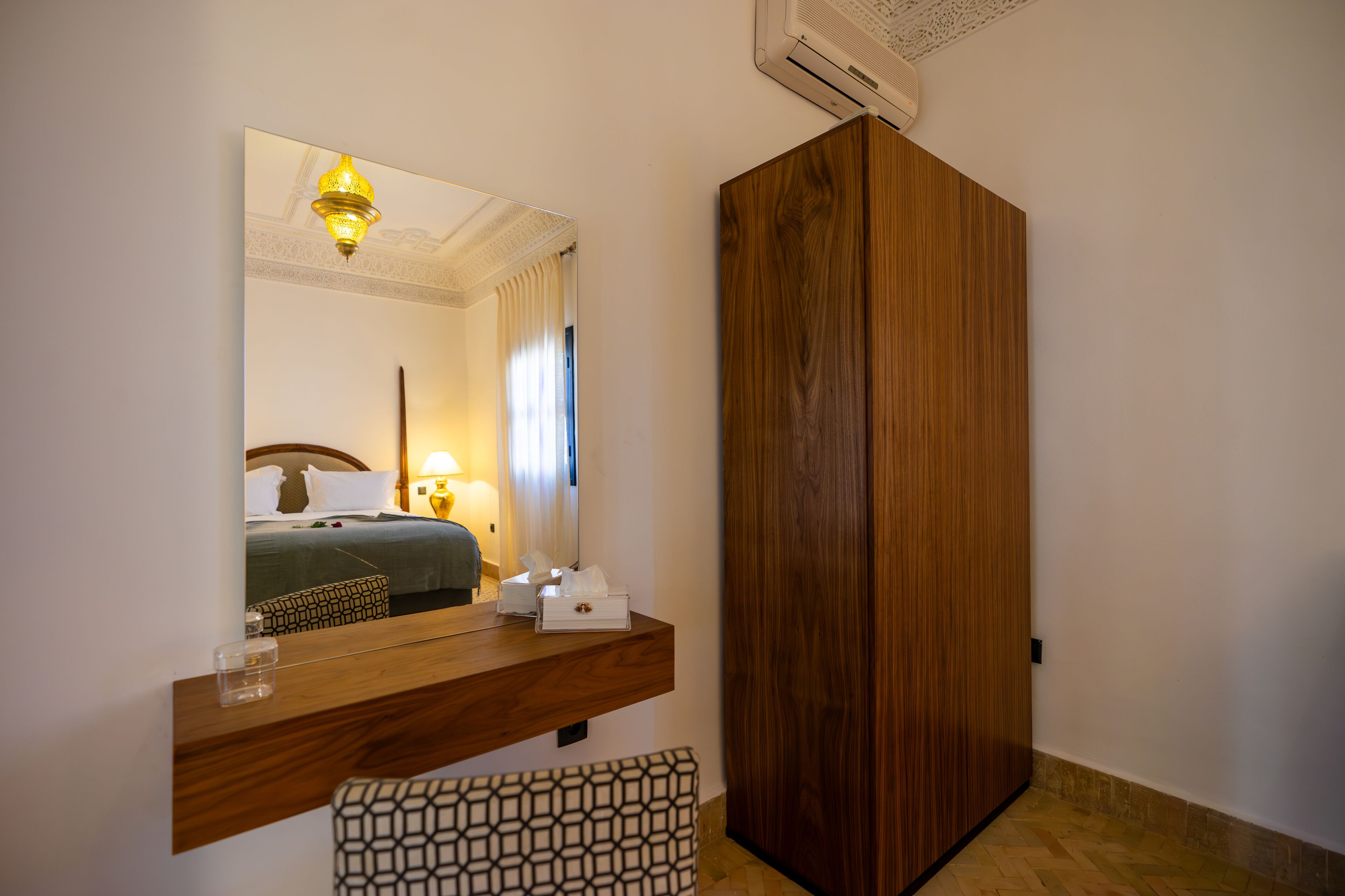 Quarto de hotel com toucador de madeira, um espelho grande, mesa de cabeceira com abajur e guarda-roupa de madeira.