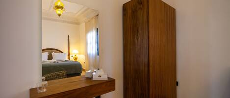 Quarto de hotel com toucador de madeira, um espelho grande, mesa de cabeceira com abajur e guarda-roupa de madeira.