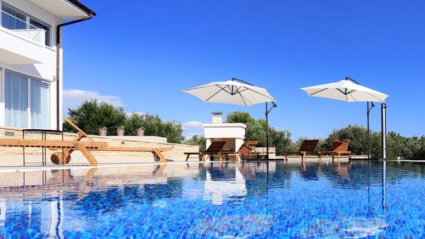 Piscina azul límpida com espreguiçadeiras e guarda-sóis, cercada por vegetação e um edifício branco.