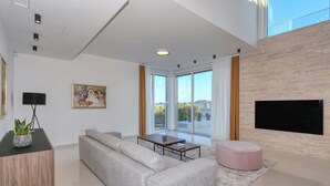 Uma sala de estar moderna com um sofá cinzento, uma mesa de centro, um banquinho rosa, uma televisão de tela plana na parede e uma grande janela com cortinas.